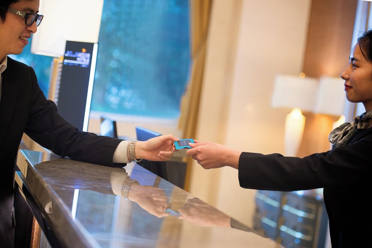 Uma área de recepção de hotel com balcão de vidro, um monitor de computador e uma janela com cortinas.