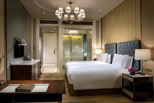 Quarto de hotel com uma cama grande, uma escrivaninha, uma cadeira, uma televisão, um candelabro e um banheiro visível através de uma porta de vidro.