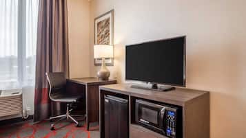 Quarto de hotel com escrivaninha, televisão de tela plana, mini-geladeira, micro-ondas, cadeira, abajur e janela com cortinas.
