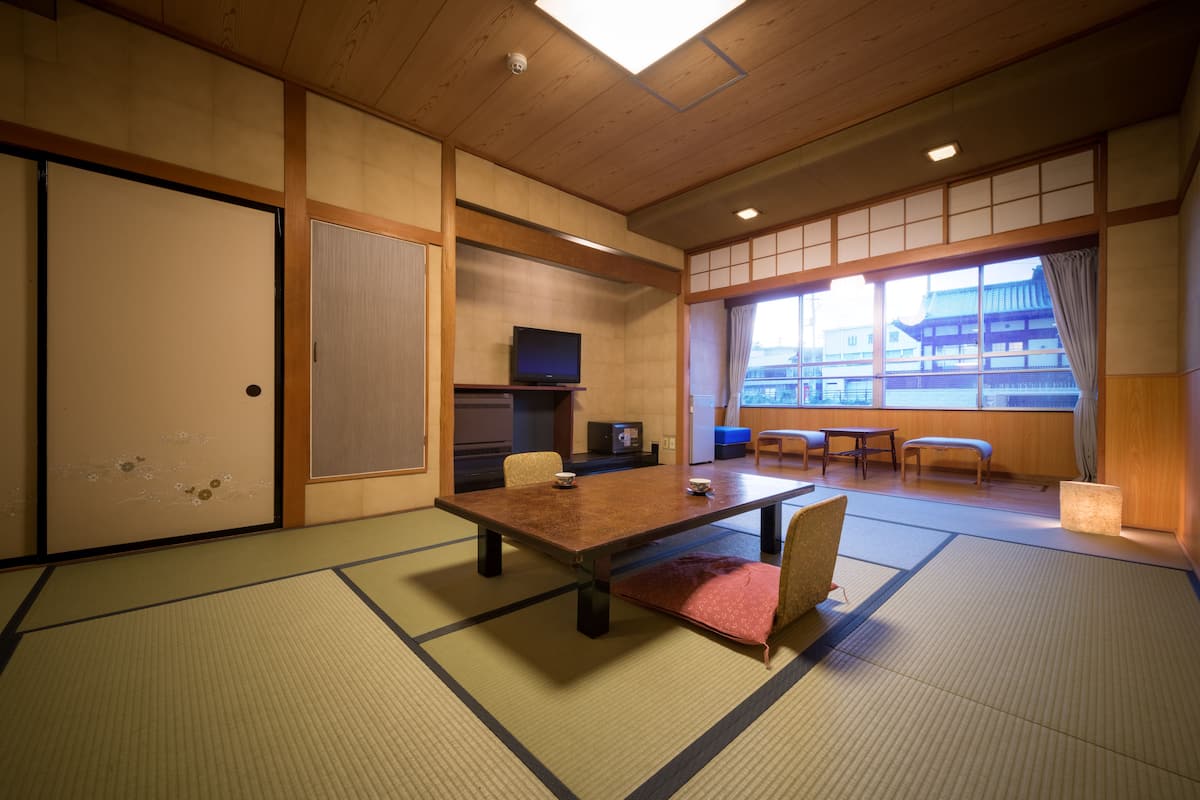 Um quarto japonês tradicional com tatames, uma mesa baixa com almofadas, uma televisão e vista para um edifício tradicional através de uma grande janela.