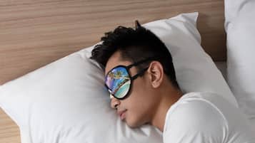 Uma pessoa deitada na cama, com roupa de cama e almofadas brancas, num quarto com cabeceira de madeira.