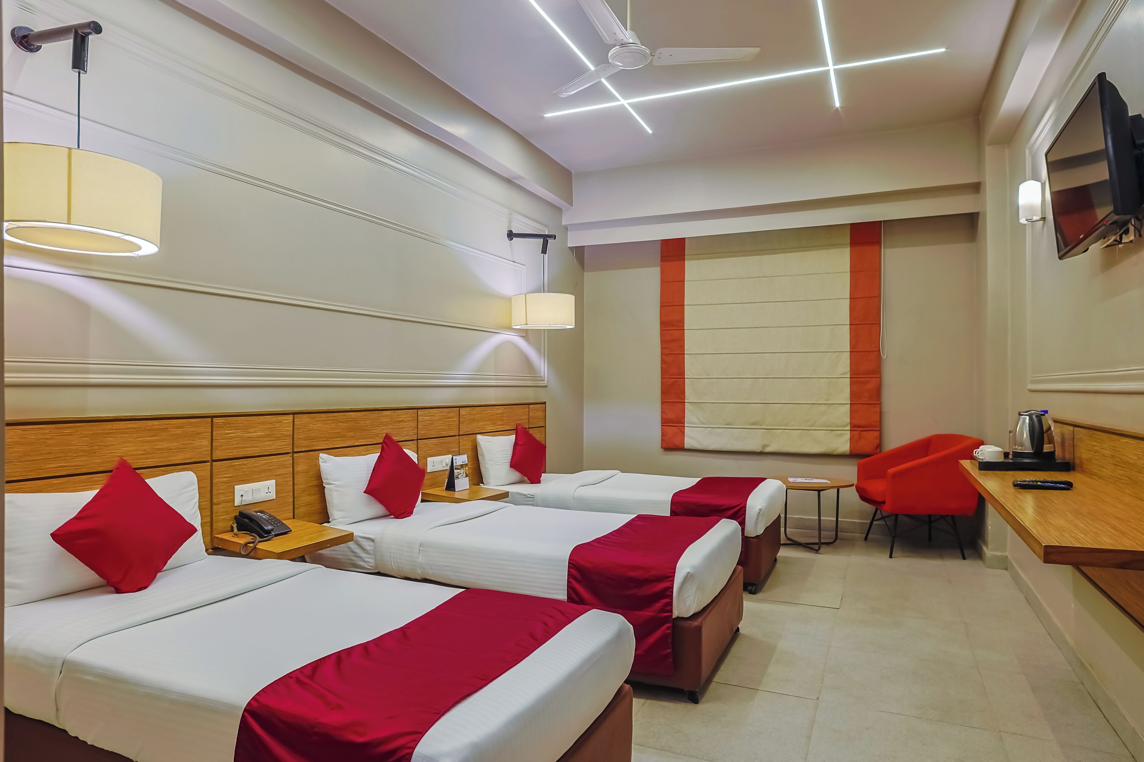 Quarto de hotel com três camas, uma escrivaninha, uma cadeira, uma televisão e uma janela com detalhe em vermelho.