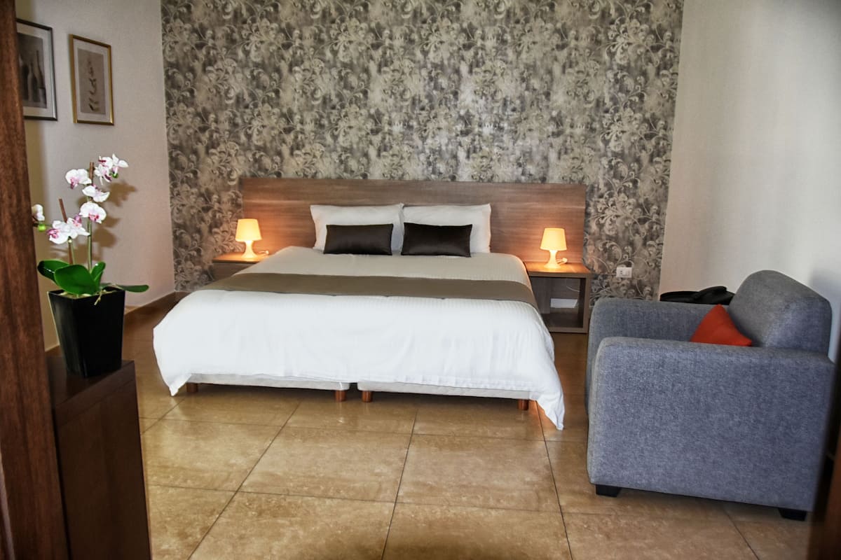 Quarto de hotel com uma cama grande, duas luminárias de cabeceira, uma poltrona cinza e um vaso de plantas.