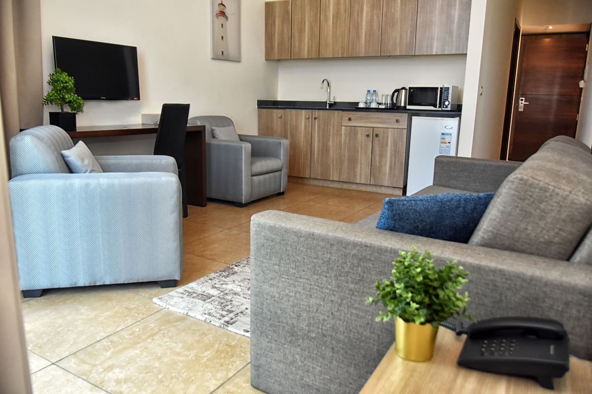 Uma sala de estar moderna com um sofá cinzento, uma poltrona cinza e uma mesa de centro com uma planta. Uma televisão de tela plana está fixada na parede, e uma cozinha é visível ao fundo.