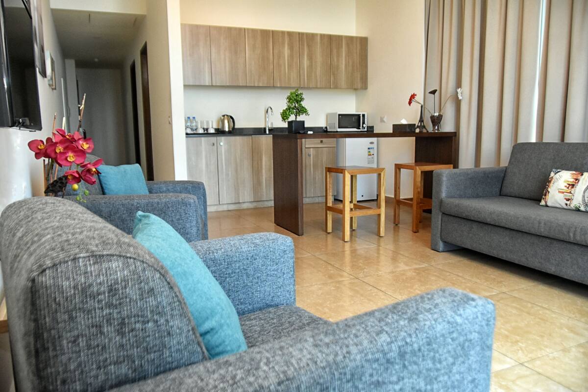Uma sala de estar moderna com sofás cinzentos, uma pequena mesa de madeira e uma área de cozinha com micro-ondas e armários.