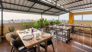 Um restaurante no terraço com várias mesas e cadeiras, plantas em vasos e vista para a paisagem urbana.