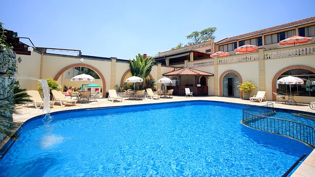 Piscina com espreguiçadeiras e guarda-sóis, cercada por um edifício com arcos e telhado em azulejos.