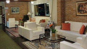 Sala de estar com sofás brancos, uma mesa de centro e um tapete estampado. Há duas luminárias pendentes, uma janela com cortinas e uma parede de tijolos com um quadro emoldurado.