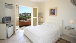 Quarto com cama, televisão, pequeno frigorífico e varanda com vista para casas.