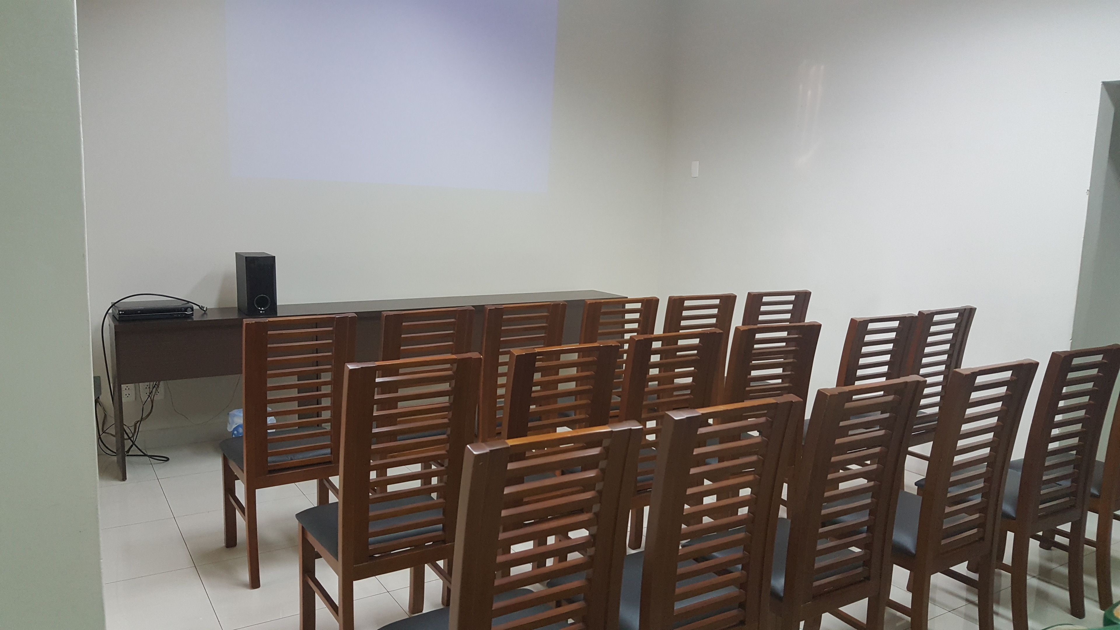 Uma sala de conferências com tela de projetor, uma mesa e várias cadeiras de madeira.