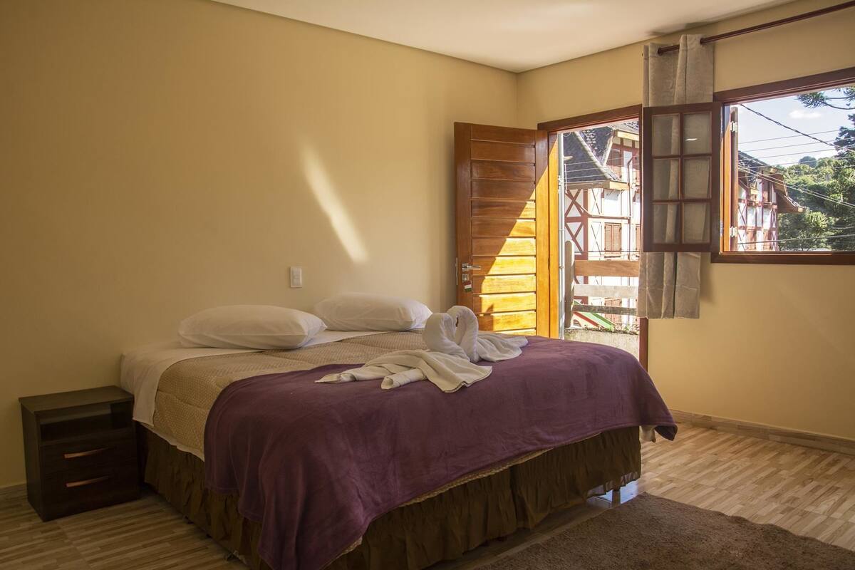 Quarto com cama, mesa de cabeceira, janela com vista para um edifício tradicional e porta de madeira.