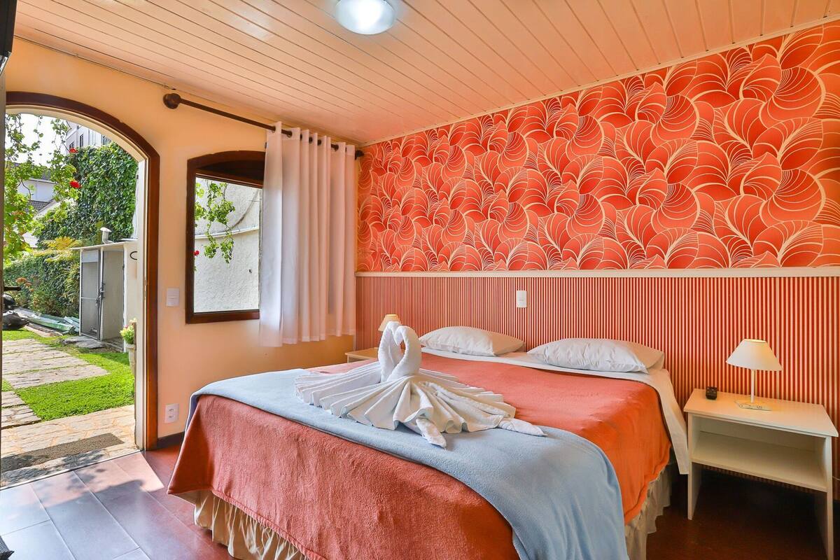 Quarto de hotel com cama, duas almofadas, mesa de cabeceira, luminária, janela com cortinas e vista para um jardim.