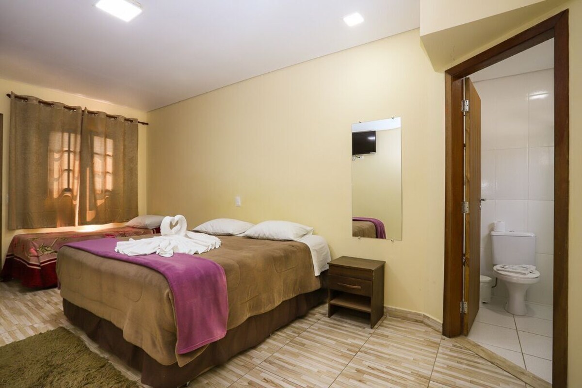 Quarto com duas camas, mesa de cabeceira, espelho e banheiro com vaso sanitário e pia.