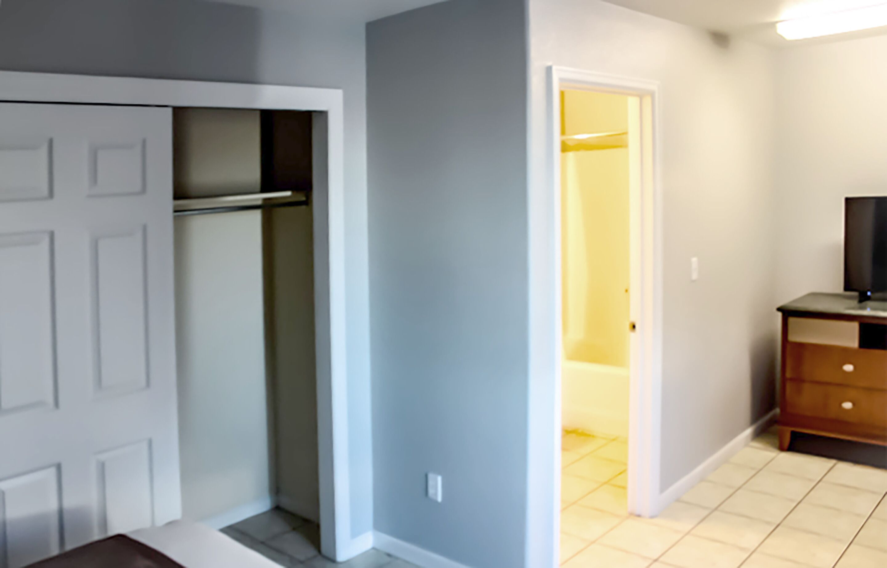 Quarto com porta fechada, armário, cômoda de madeira e televisão.