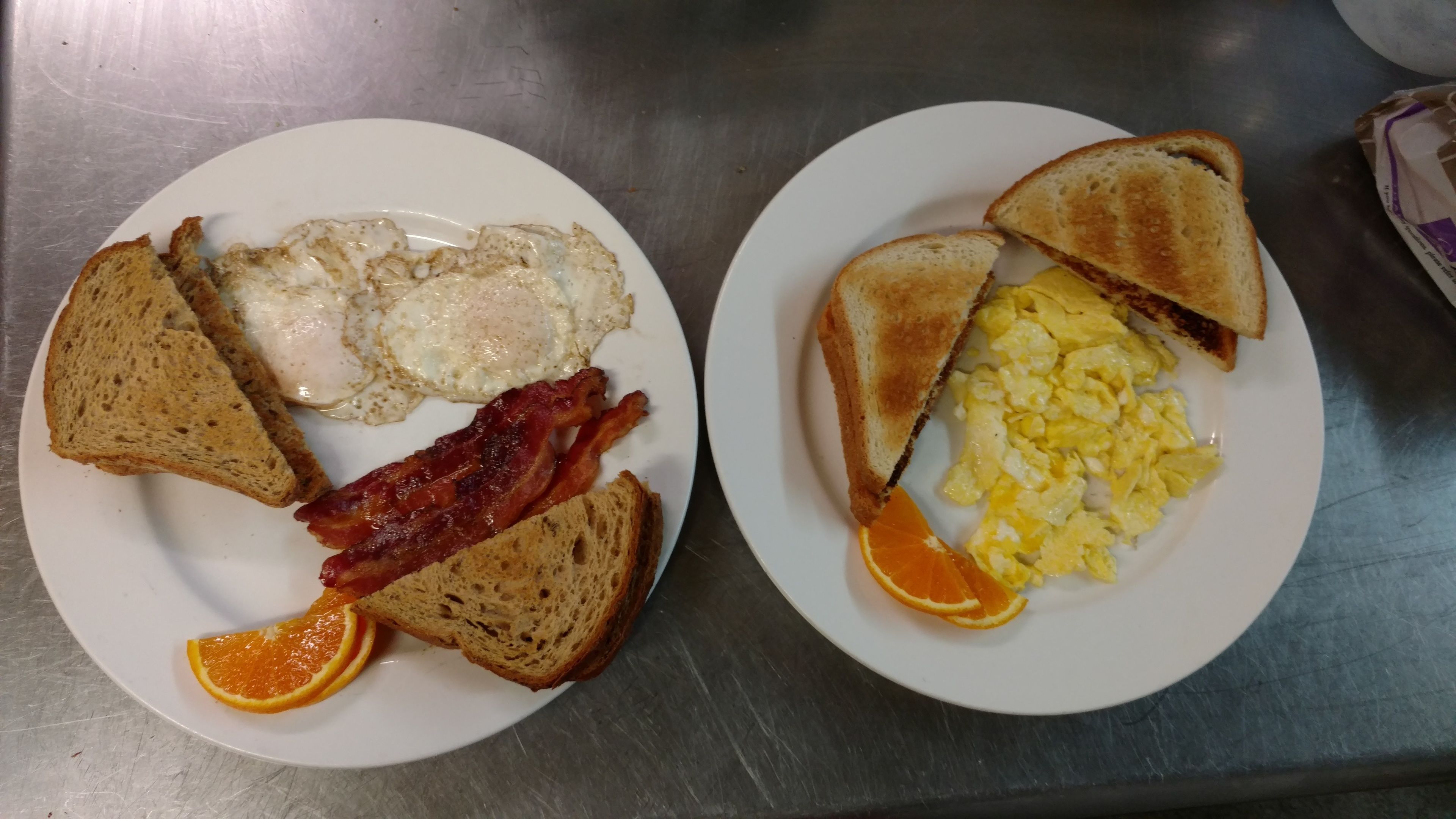 Duas travessas de comida de café da manhã, incluindo torradas, ovos e bacon, sobre uma mesa de aço inoxidável.
