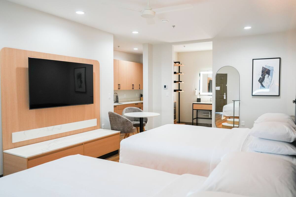 Quarto de hotel com cama, televisão na parede, uma mesinha e um espaço de estar com um banco.