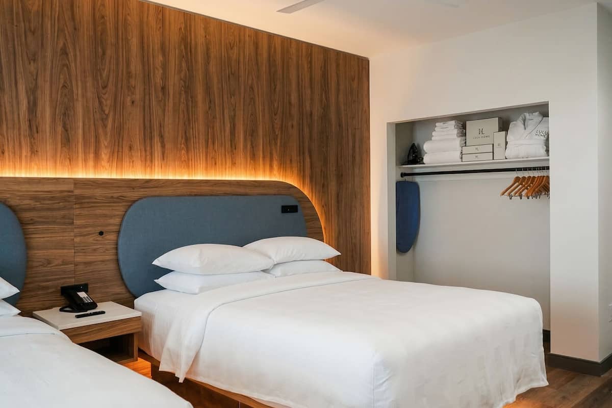Quarto de hotel com cabeceira de madeira, duas camas, um guarda-roupa com roupas penduradas e uma prateleira com toalhas dobradas.