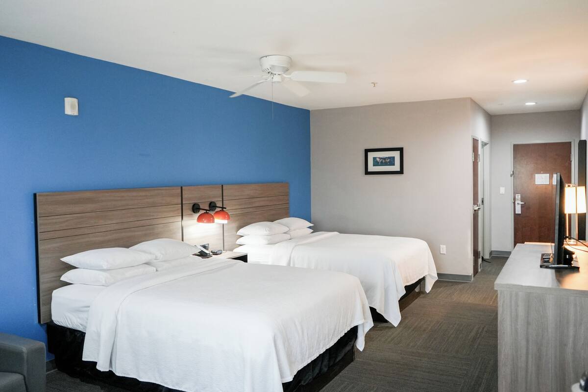 Quarto de hotel com duas camas, um ventilador de teto, uma mesa com abajur e uma porta que dá para outro quarto.