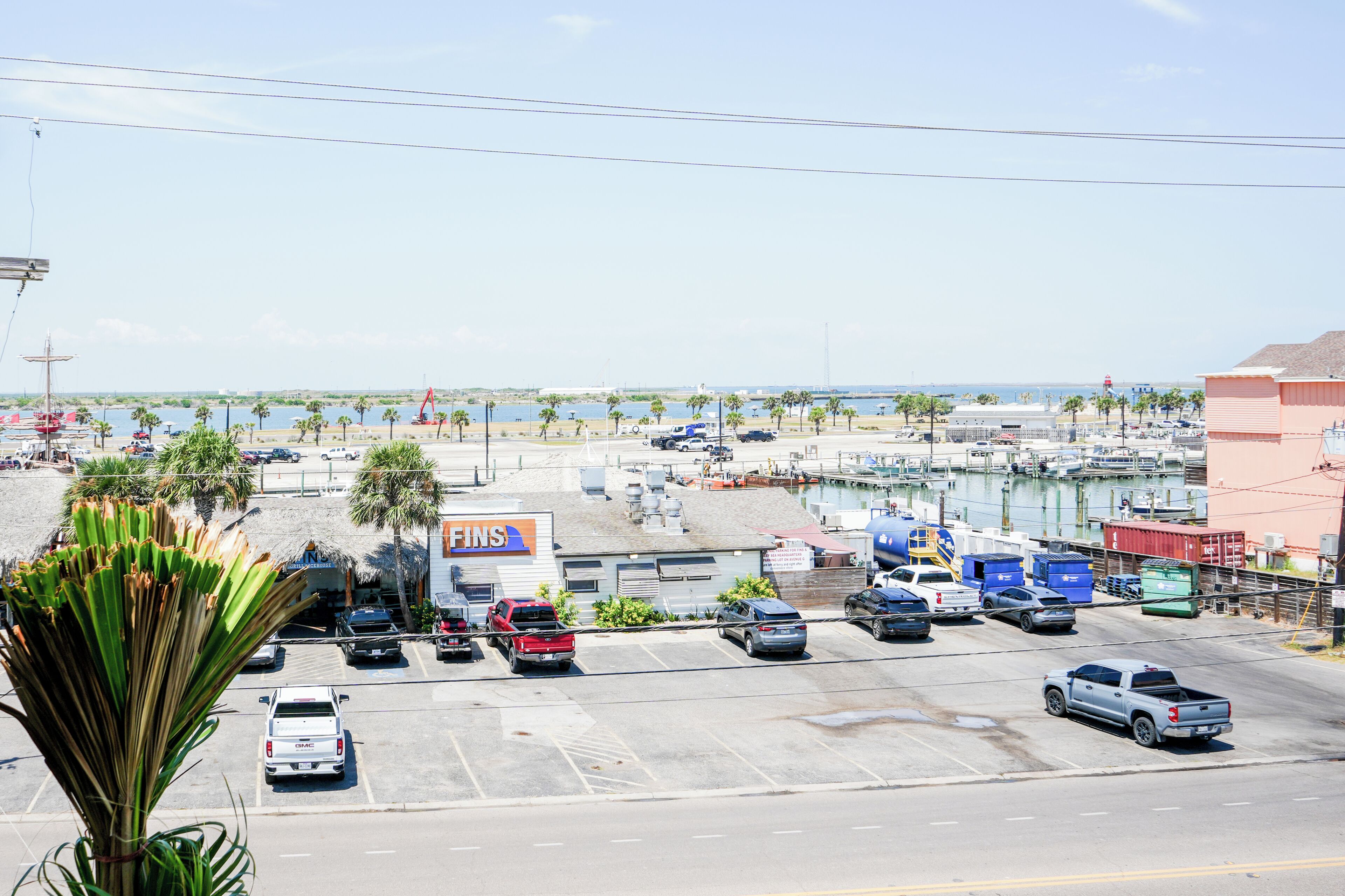 Um estacionamento com vários veículos, um edifício com uma placa e uma praia ao fundo.