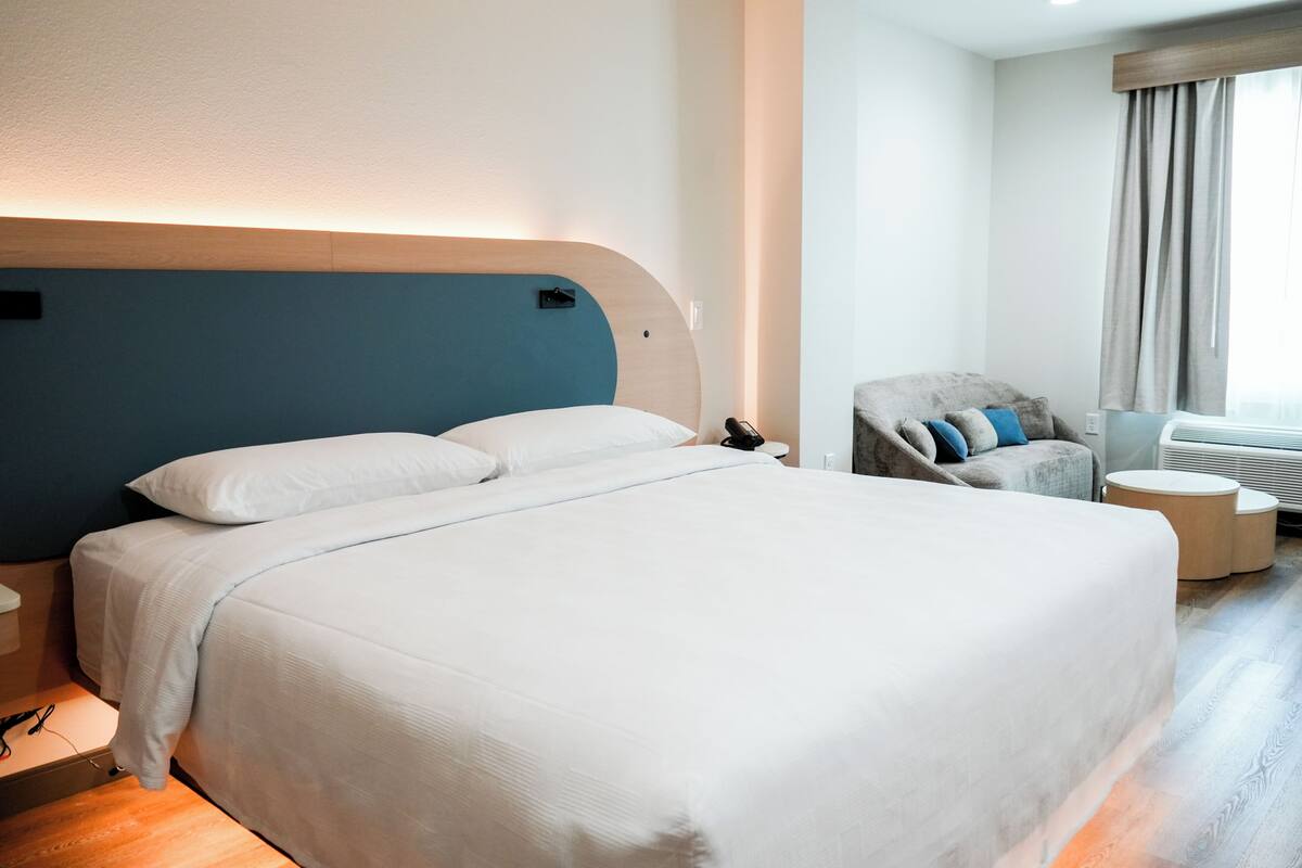Quarto de hotel moderno com uma cama grande, uma cadeira, uma mesinha e um aparelho de ar condicionado.