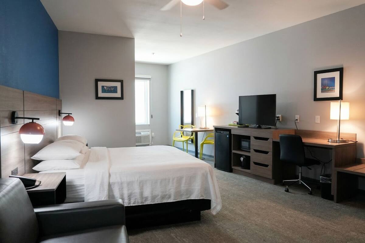 Quarto de hotel com cama, escrivaninha, cadeira, televisão e duas luminárias.