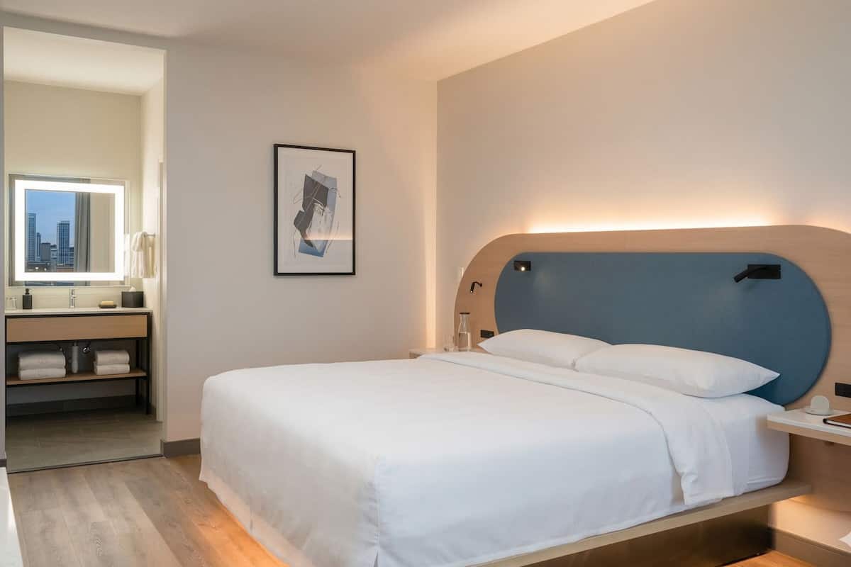 Quarto de hotel moderno com uma cama grande, uma mesa de cabeceira, um espelho e um quadro na parede.