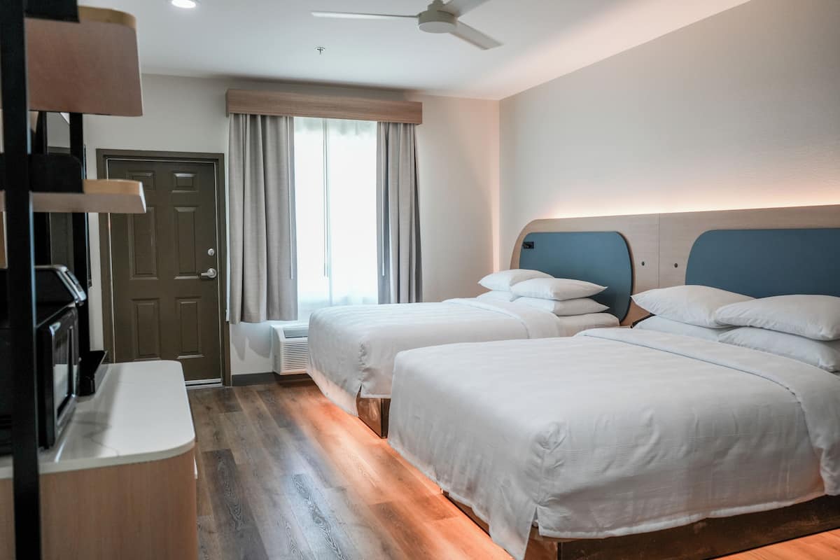 Quarto de hotel com duas camas, uma escrivaninha, um ventilador de teto e uma janela com cortinas.