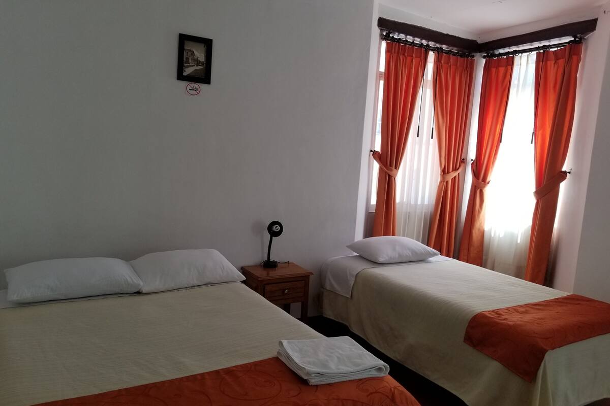 Quarto de hotel com duas camas, uma mesinha de cabeceira, uma luminária e uma janela com cortinas laranja.