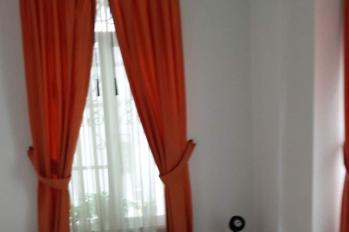 Uma cama com almofadas brancas e um edredom vermelho, uma mesa de cabeceira com uma luminária, e cortinas laranja.