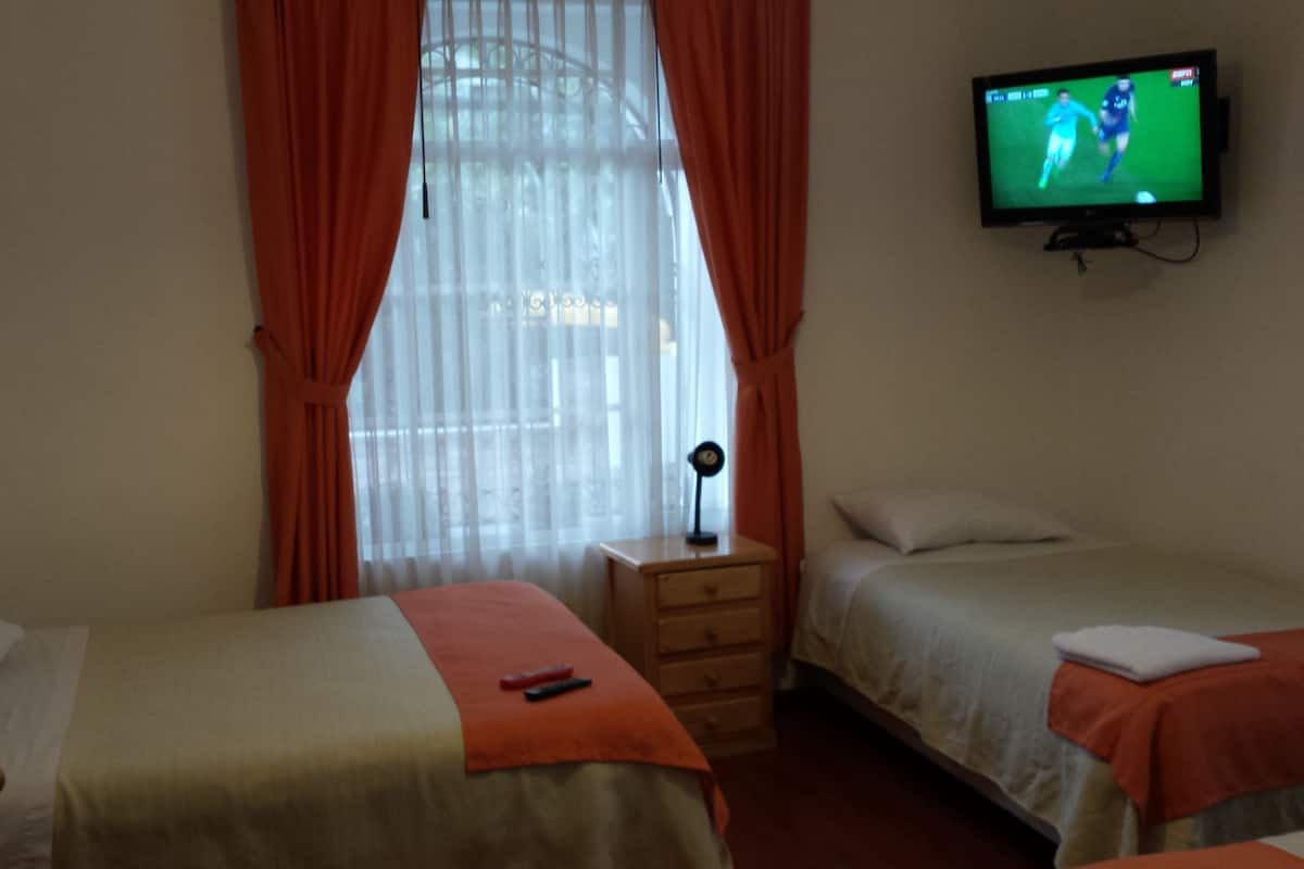 Quarto de hotel com duas camas, uma mesinha de cabeceira, televisão e uma janela com cortinas.
