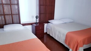 Duas camas de solteiro com roupa de cama branca e laranja, uma mesinha de cabeceira de madeira e uma luminária fixada na parede.