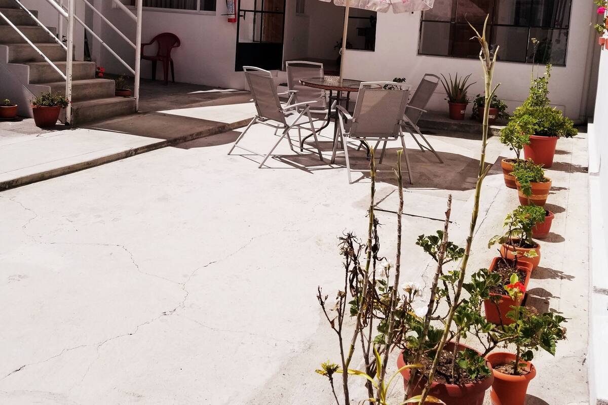 Uma área de varanda externa com mesa e cadeiras, plantas em vasos e uma escadaria que leva a um andar superior.
