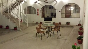 Um espaço interno amplo com uma mesa de vidro central e cadeiras, cercado por plantas em vasos. Uma escada branca com grades de metal é visível à esquerda, levando a um andar superior.