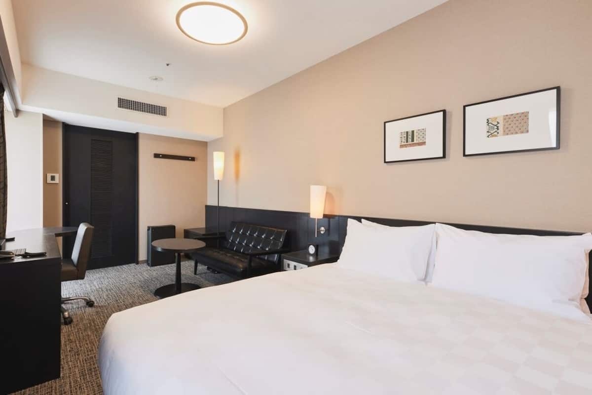 Quarto de hotel com cama, escrivaninha, cadeira, mesa pequena e duas fotos emolduradas na parede.