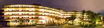 Um hotel com vários andares e fachada curva, com várias varandas e uma área de entrada bem iluminada com palmeiras.
