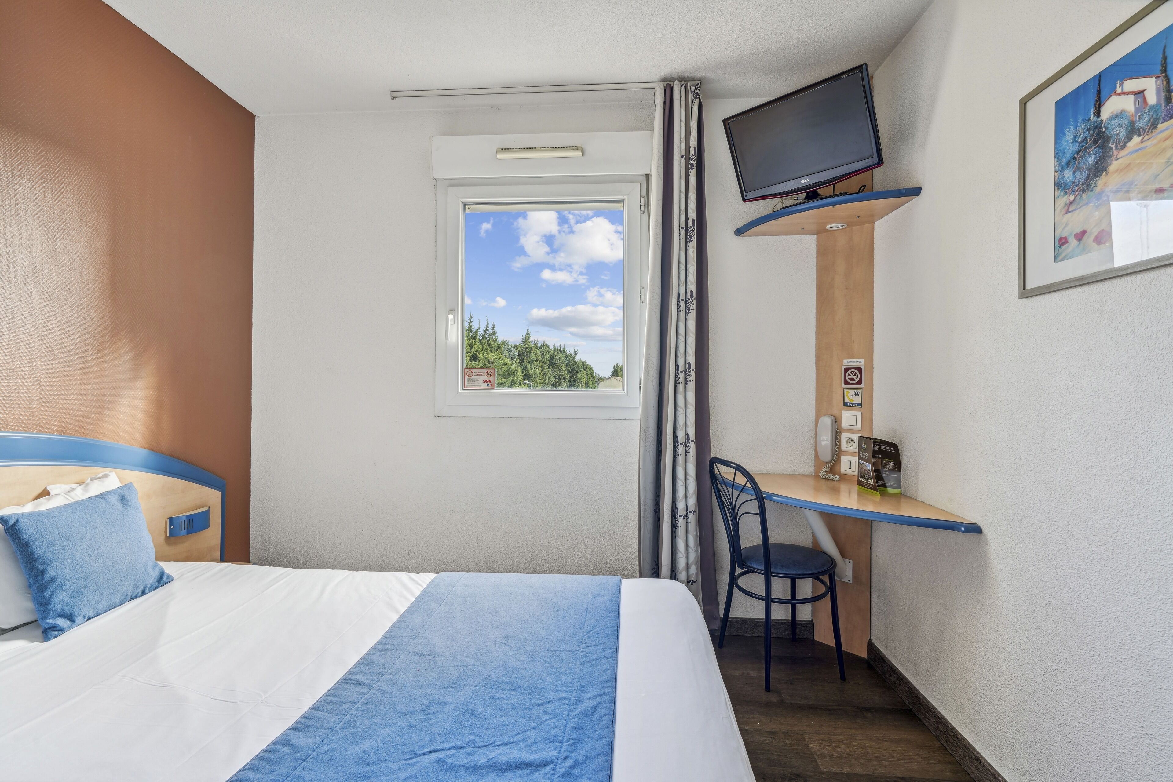 Quarto de hotel com cama, escrivaninha, cadeira, televisão e janela com vista para árvores.