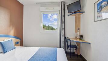 Quarto de hotel com cama, escrivaninha, cadeira, televisão e janela com vista para árvores.