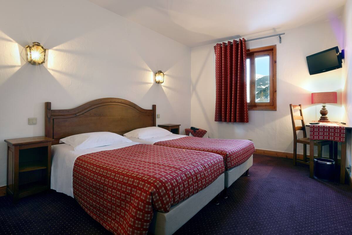 Quarto de hotel com duas camas, televisão, secretária com candeeiro e janela com cortinas.