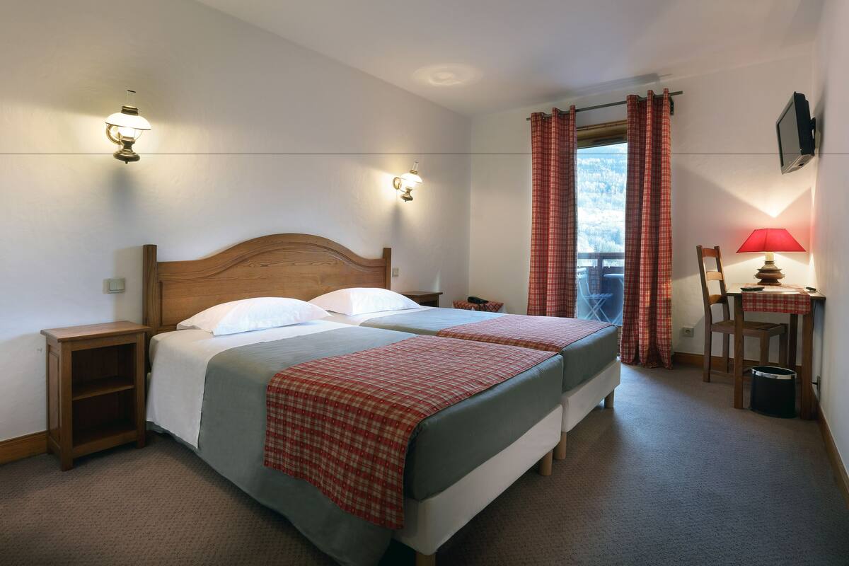 Quarto de hotel com duas camas, uma mesa de escritório com cadeira, televisão e janela com cortinas.