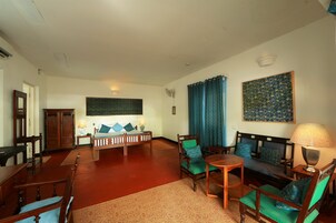 Uma sala de estar ampla com um conjunto de sofá de madeira, uma mesa de centro redonda e uma mesa de jantar com cadeiras. Há uma janela grande com cortinas azuis, um ventilador fixado na parede e um objeto decorativo na parede.