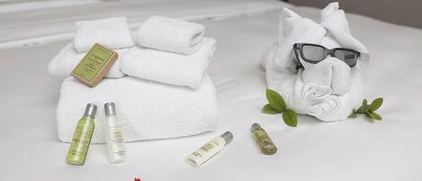 Cama bem arrumada com lençóis brancos, toalha enrolada e duas embalagens de loção.