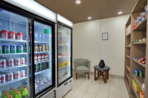 Quarto com geladeira repleta de bebidas, uma cadeira e prateleiras com petiscos.