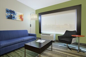 Quarto de hotel moderno com sofá azul, uma mesa pequena, uma poltrona preta, uma mesa redonda e uma grande janela com vista para a cidade.