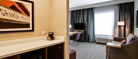 Quarto de hotel com cozinha compacta, TV, escrivaninha, sofá e uma janela grande com cortinas.