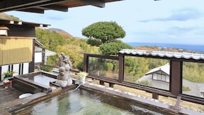 Uma onsen japonesa tradicional com um deque de madeira, nascente termal de pedra e vista para o oceano e colinas.