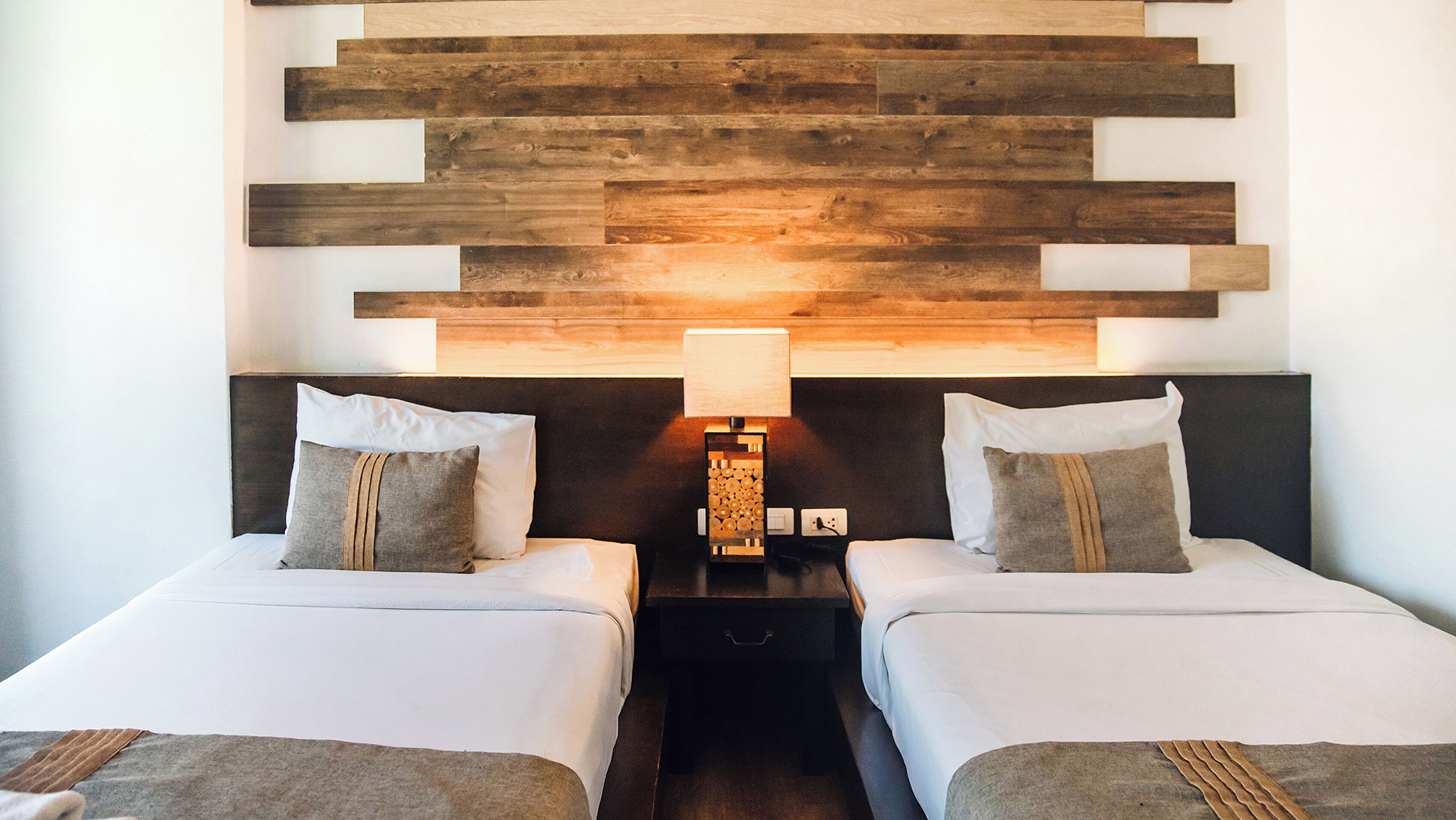 Quarto de hotel com duas camas, cabeceira de madeira, mesa de cabeceira com abajur e luminária na parede.