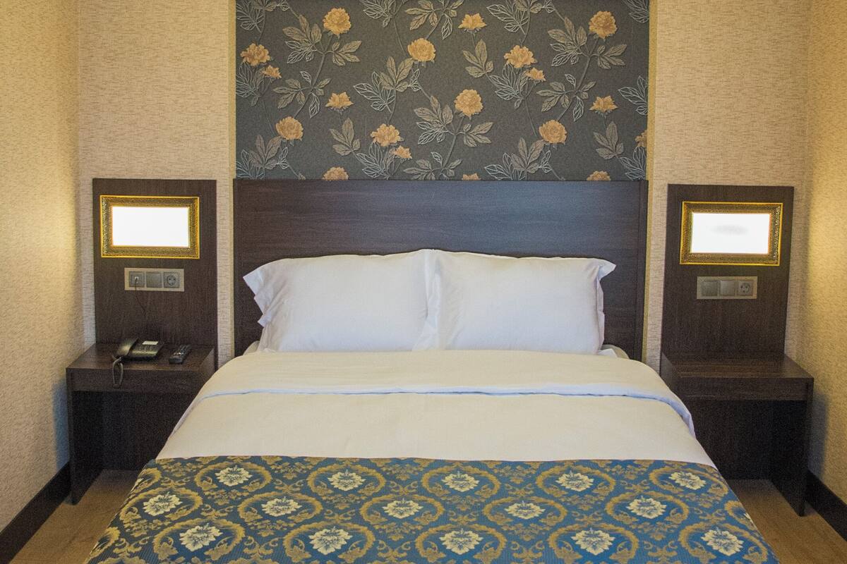 Quarto de hotel com uma cama grande, duas mesas de cabeceira com luminárias, um painel de cabeceira com padrão floral e uma luz no teto.