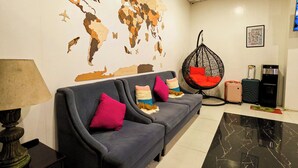 Quarto com um mural de mapa mundi, um sofá cinzento com almofadas coloridas, uma cadeira suspensa, uma máquina de café e piso ladrilhado.