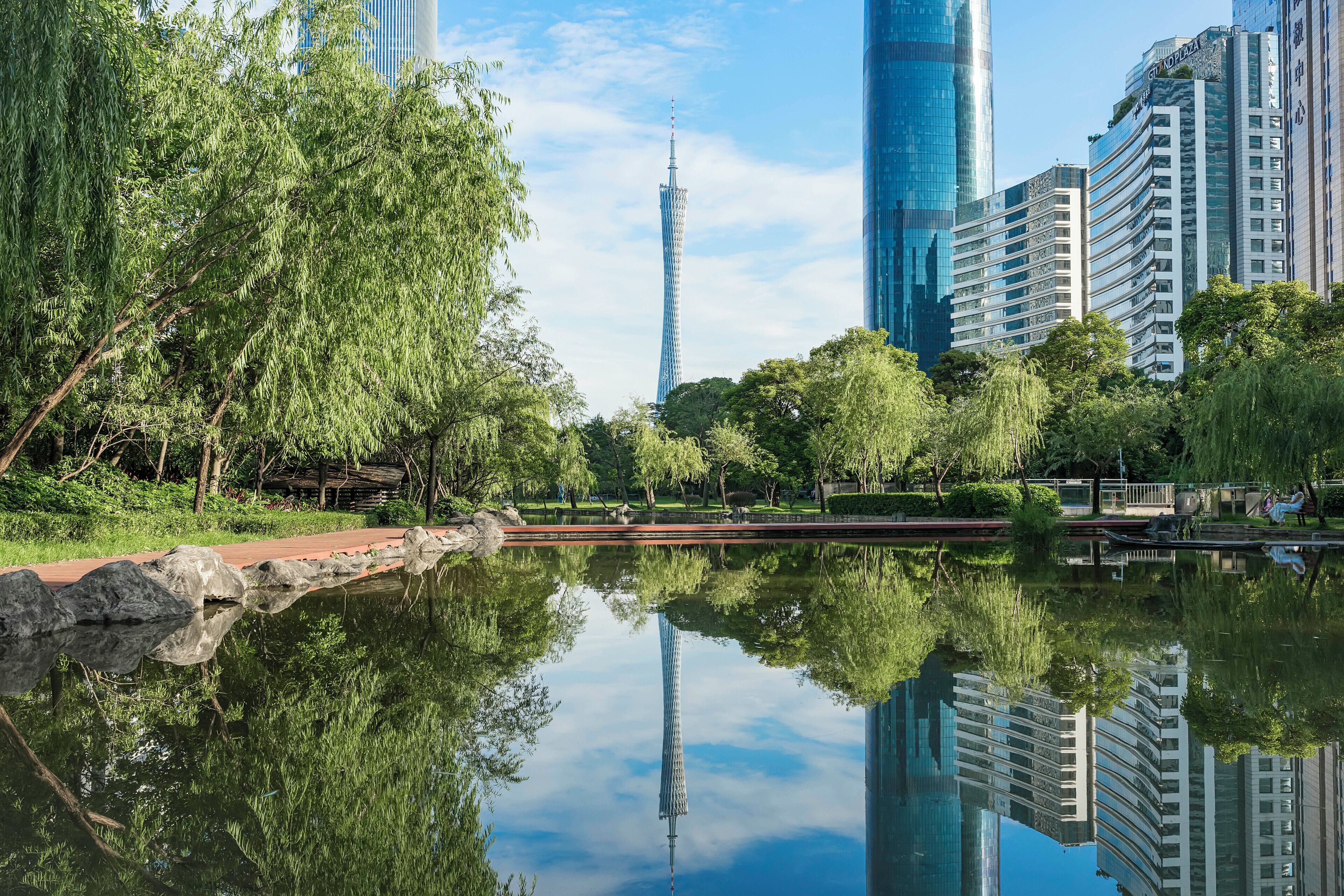 Um parque com um lago reflexivo, árvores e edifícios modernos, incluindo uma torre alta.