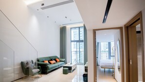Quarto de hotel moderno com um sofá verde, uma mesa pequena, uma divisória de vidro e vista para a cidade através de grandes janelas.